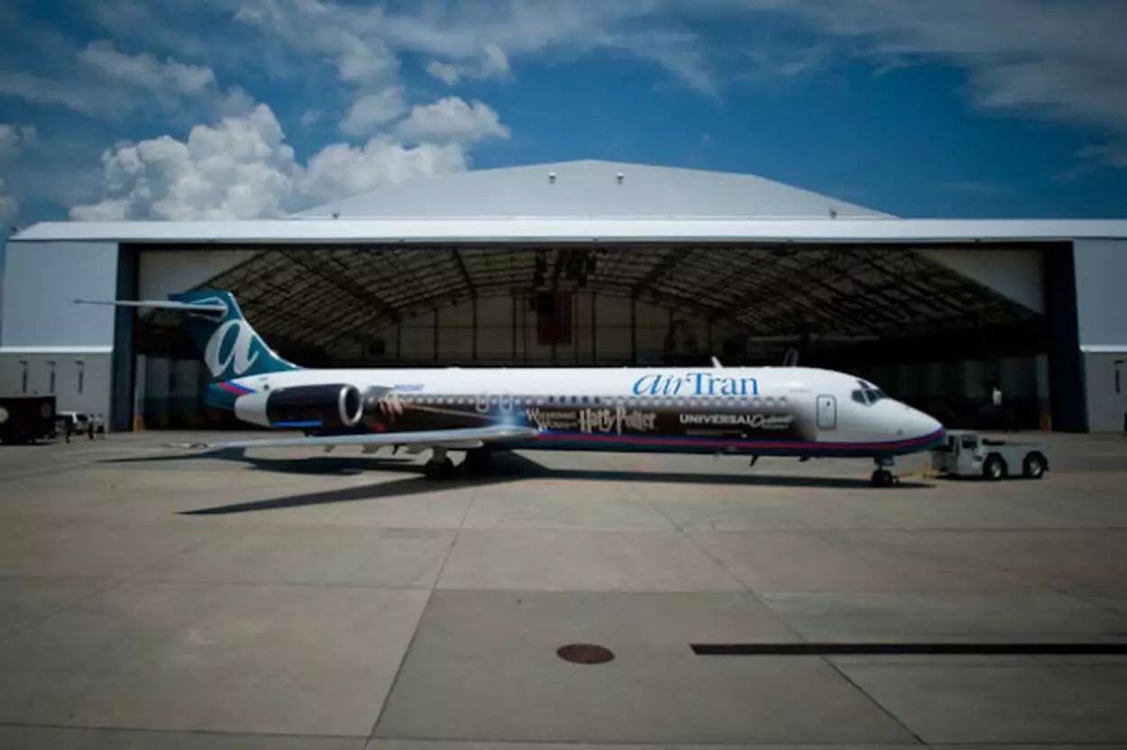 ဟယ်ရီပေါ်တာ။ AirTran Airways မှ Wizarding World of Harry Potter - Boeing 717-231 လက်တစ်ဖက်က မှော်ဓားဆွဲနဲ့ မှော်ဒြပ်တစ်ခုရဲ့ နာမည်ကို ဆုပ်ကိုင်ထားတဲ့ လက်တစ်ဖက်ကို ဆွဲထားတယ်။